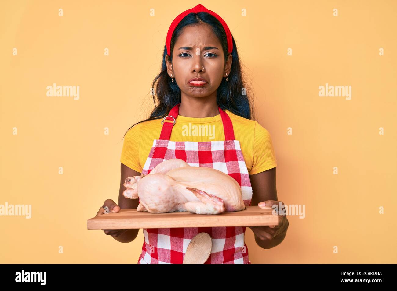 Young indian girl wearing cook apron and holding chicken depressed and ...