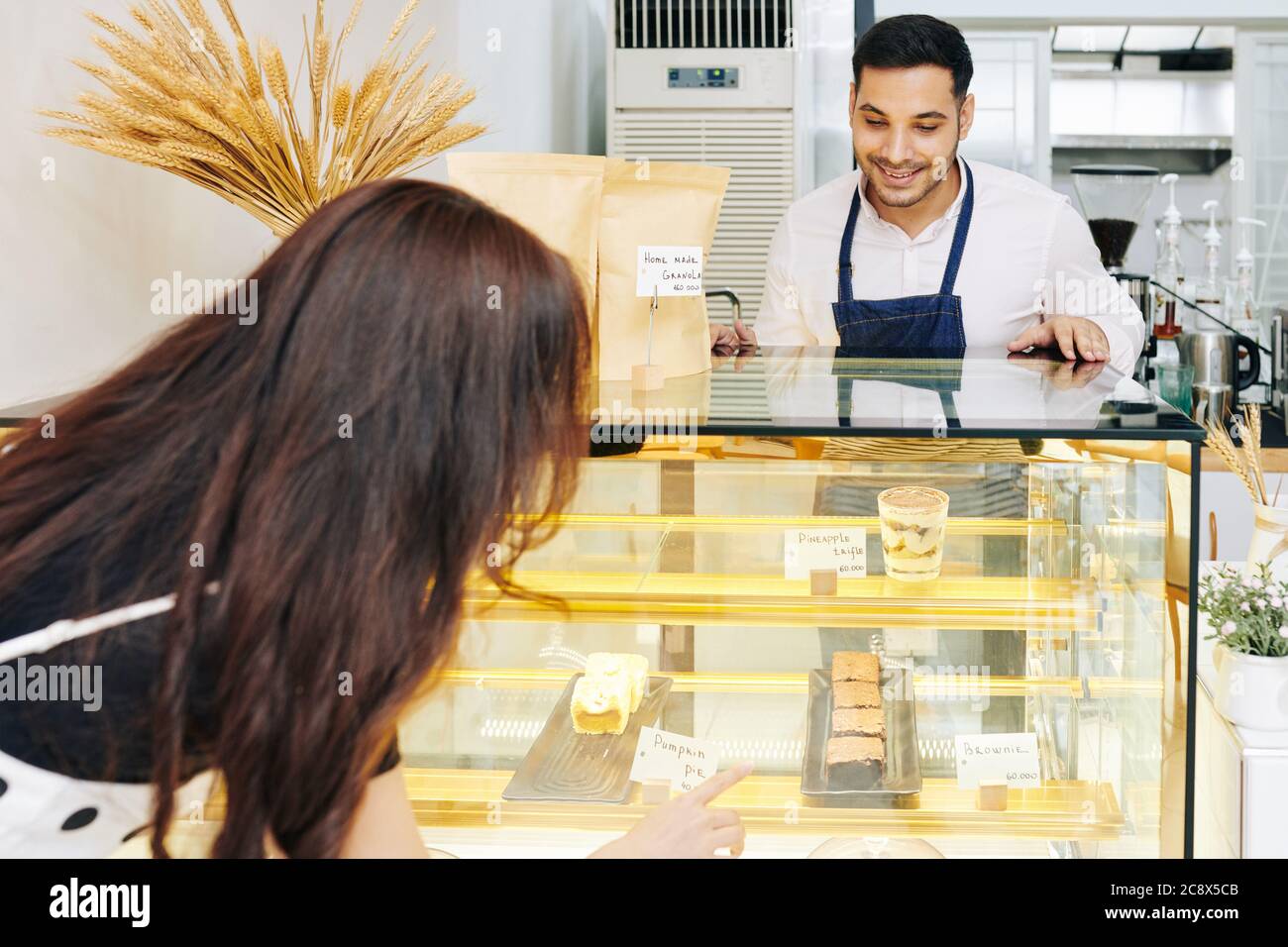 Young woman choosing dessert in showcase of bakery and asking waiter to ...