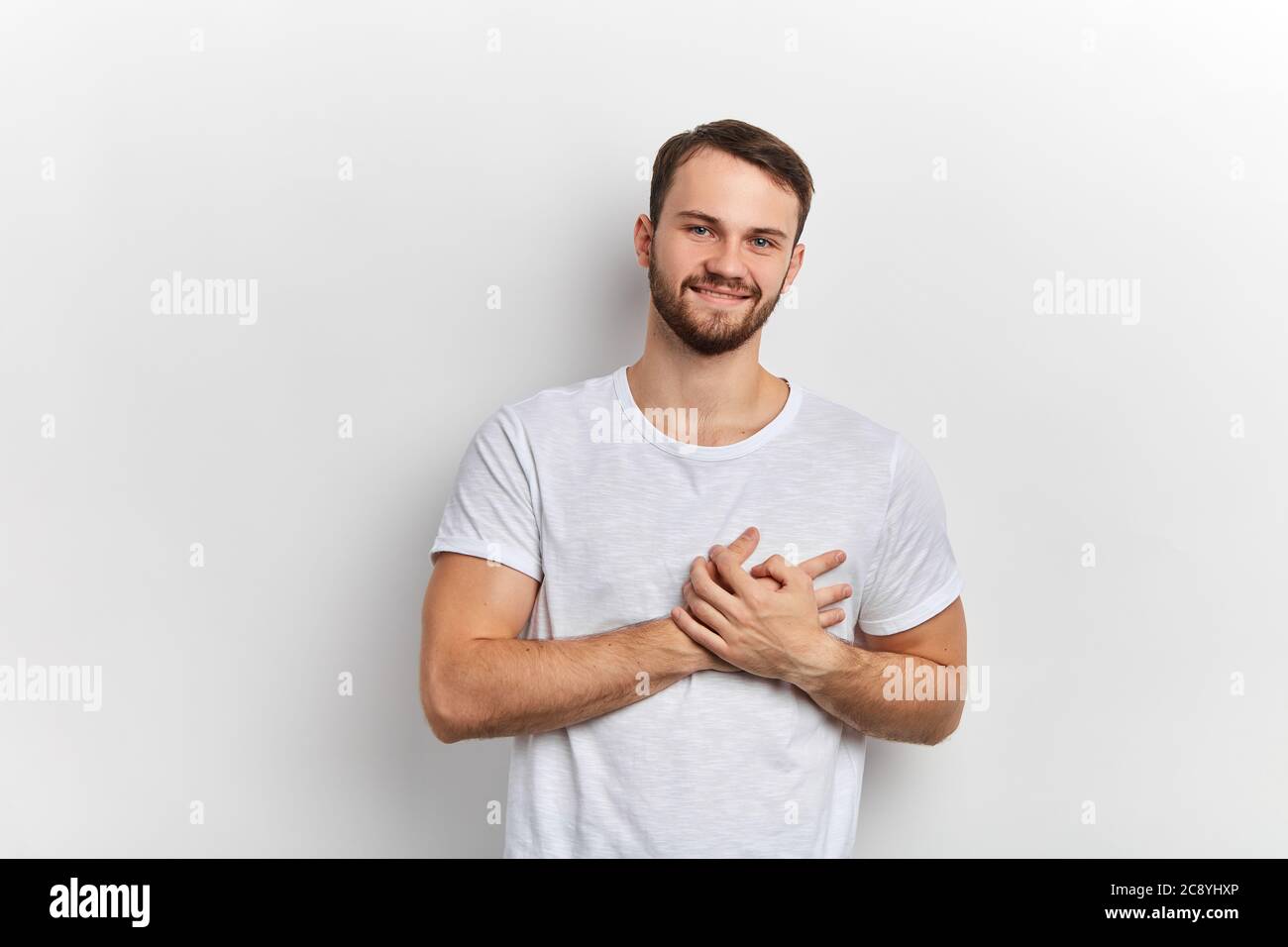 cheerful young man has friendly expression, keeps hands on heart ...