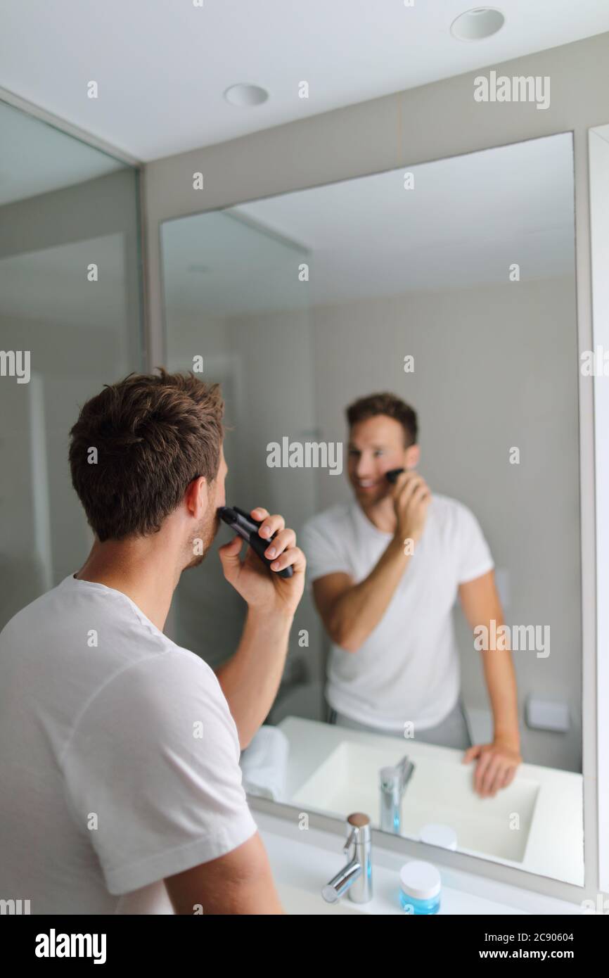 Man shaving using electric shaver trimming his beard in home bathroom- morning grooming routine ...