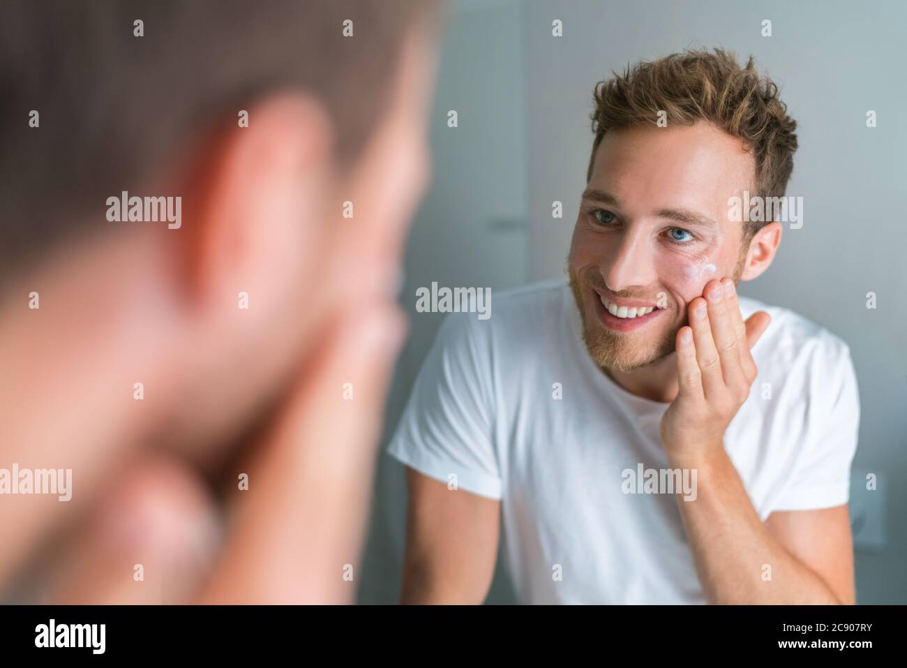 Handsome young man putting facial treatment face cream on clean skin in ...