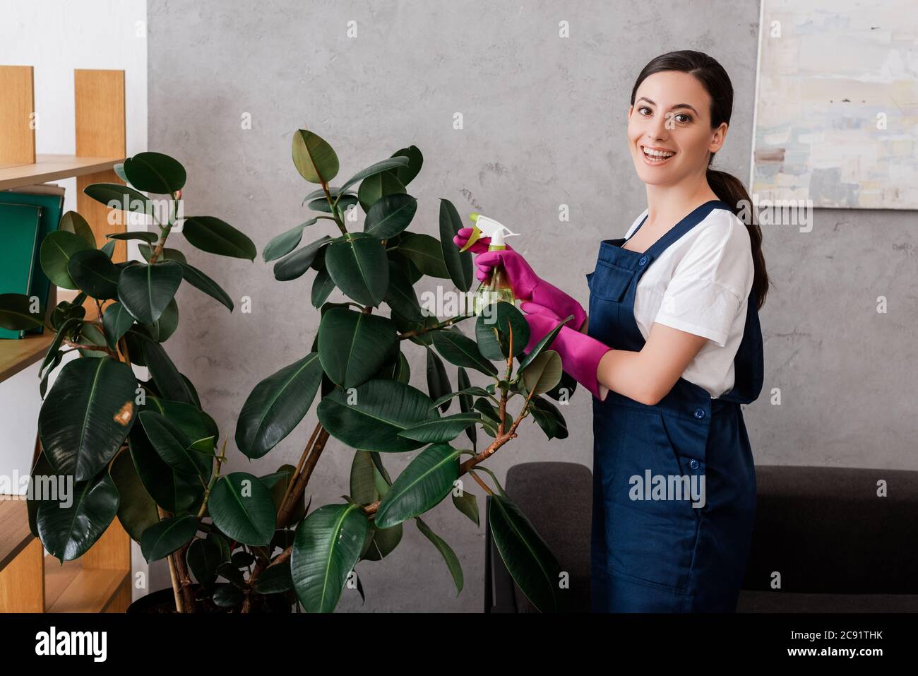 Side view of smiling cleaner holding spray bottle near plant at home ...