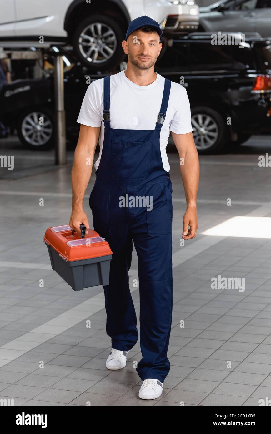 handsome mechanic in uniform and cap holding toolbox in service station ...