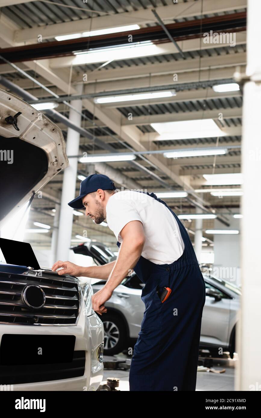 Auto mechanic leaning forward and typing on laptop on open hood of car ...