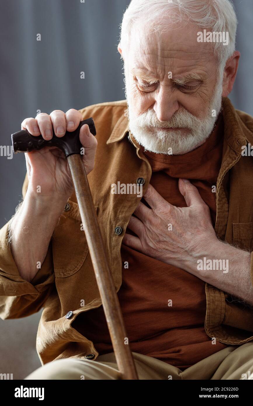 lonely senior man sitting with walking stick and touching chest while ...