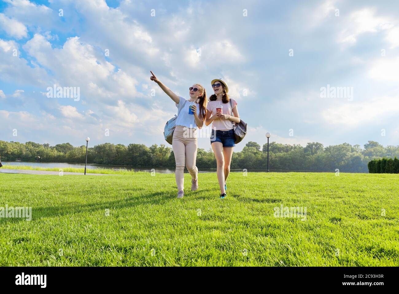 Two teenage girls walking together on the lawn with drinks on sunny ...