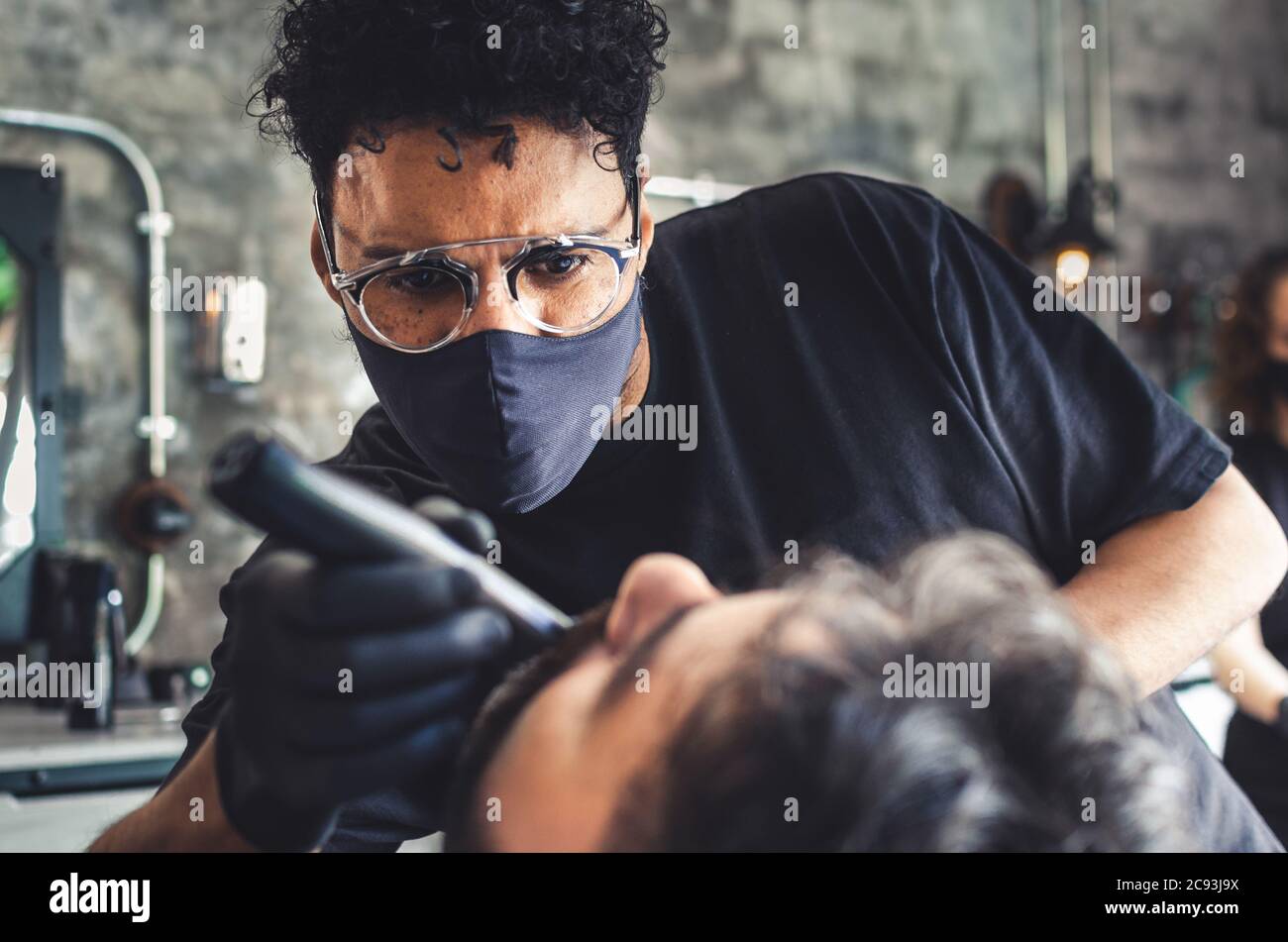 Barber with protection mask and black gloves trimming a beard with an ...