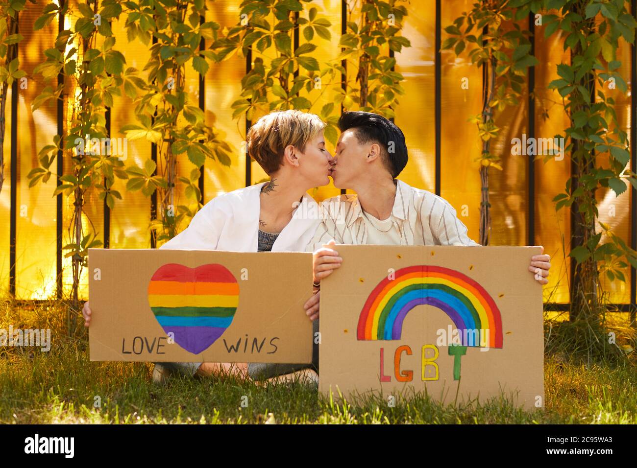 Two Lesbians Kissing And Holding Posters While Sitting On The Grass They Promoting Their Love