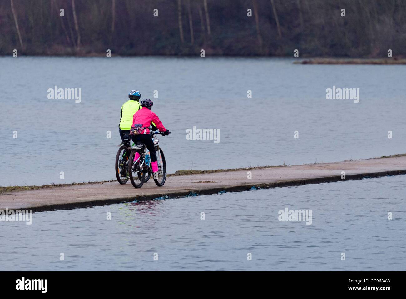 2 cyclists cycling & riding bikes (cycle ride) on narrow causeway ...