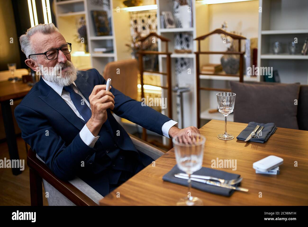Serious grey-haired elderly man in tuxedo smoke IQOS in restaurant ...