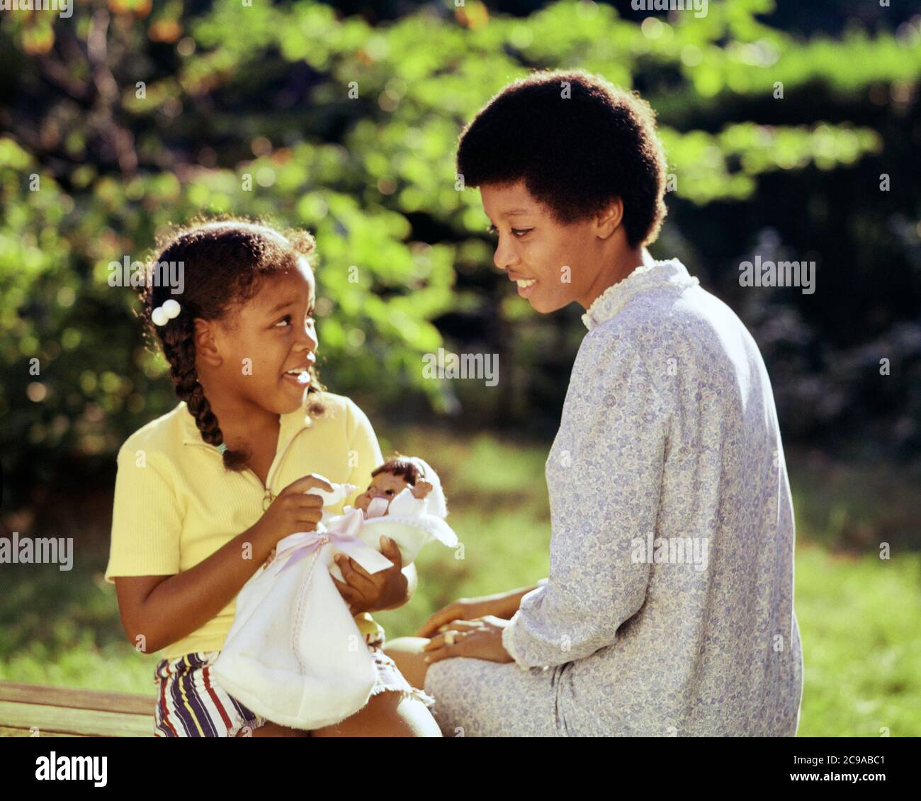 1970s AFRICAN-AMERICAN WOMAN MOTHER SITTING IN BACKYARD WITH GIRL DAUGHTER PLAYING WITH TOY BABY ...