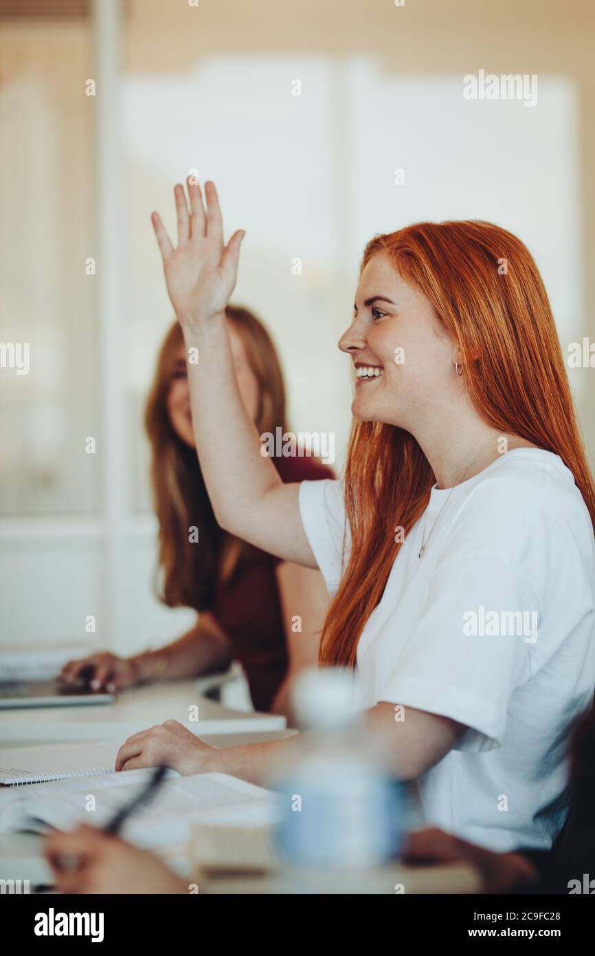 Smiling female student sitting in the class and raising hand up to ask ...