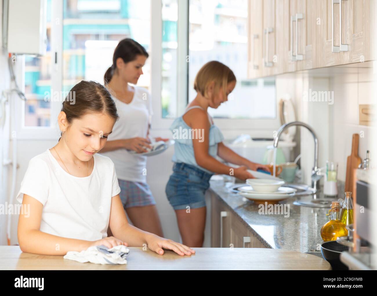 Little smiling girl cleaning kitchen surfaces at home while her mother ...