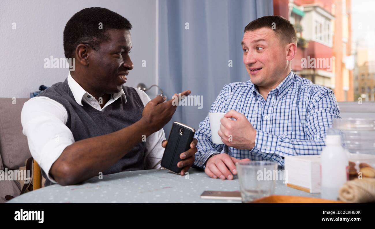 Two cheerful men enjoying conversation over cup of tea at home table ...