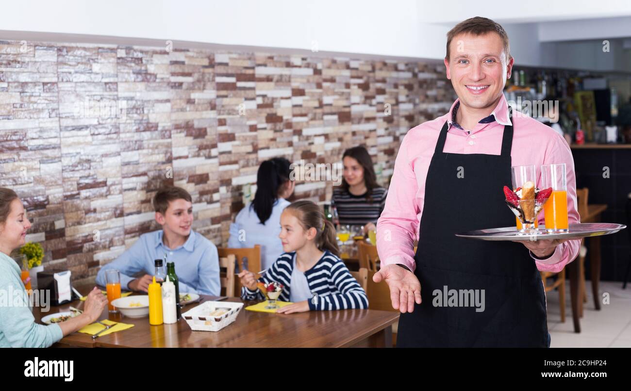 Smiling young waiter serving mother with two teenage children in cafe ...