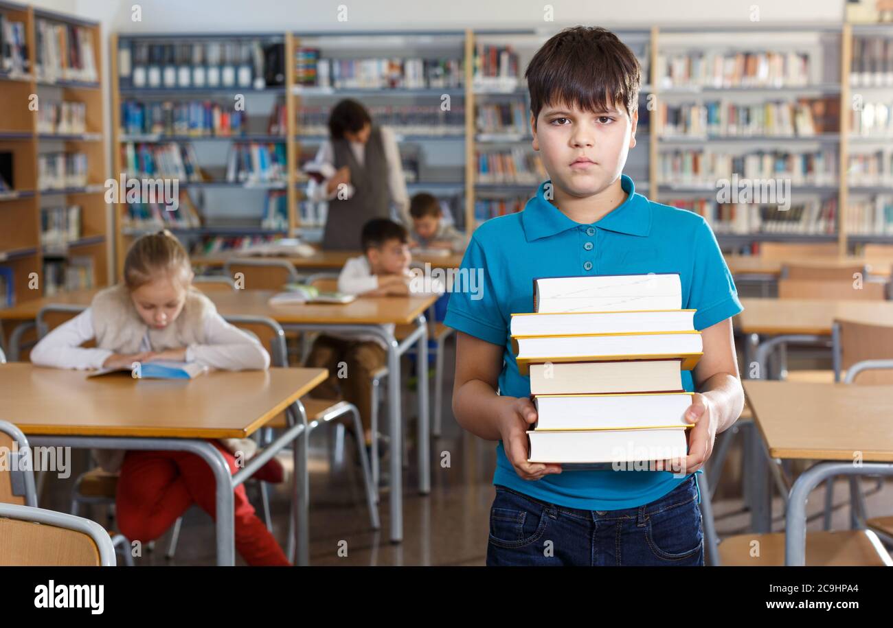 Upset tween boy standing in school library with pile of books in hands ...