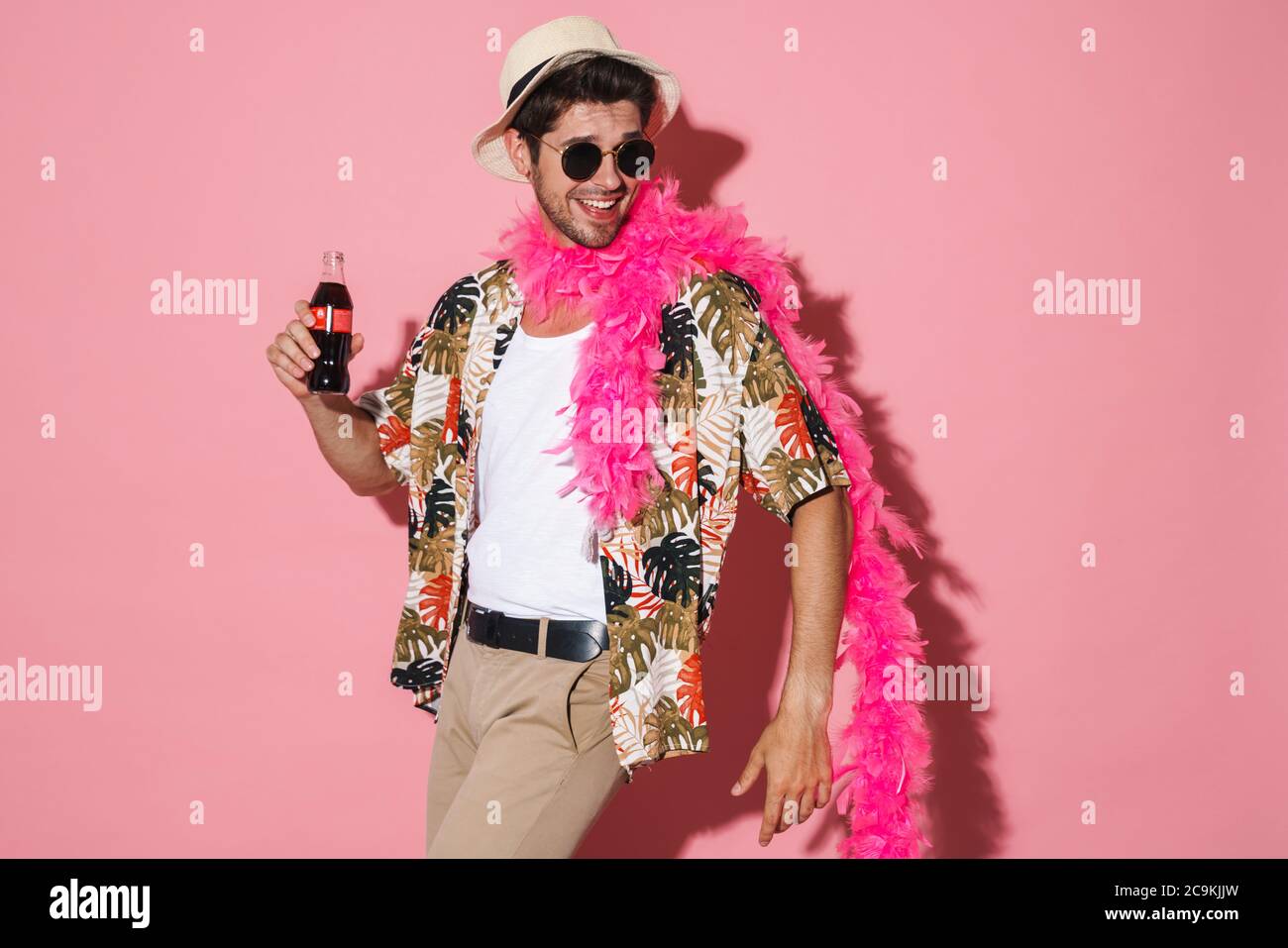 Portrait of cheerful young man wearing boa dancing while drinking soda ...