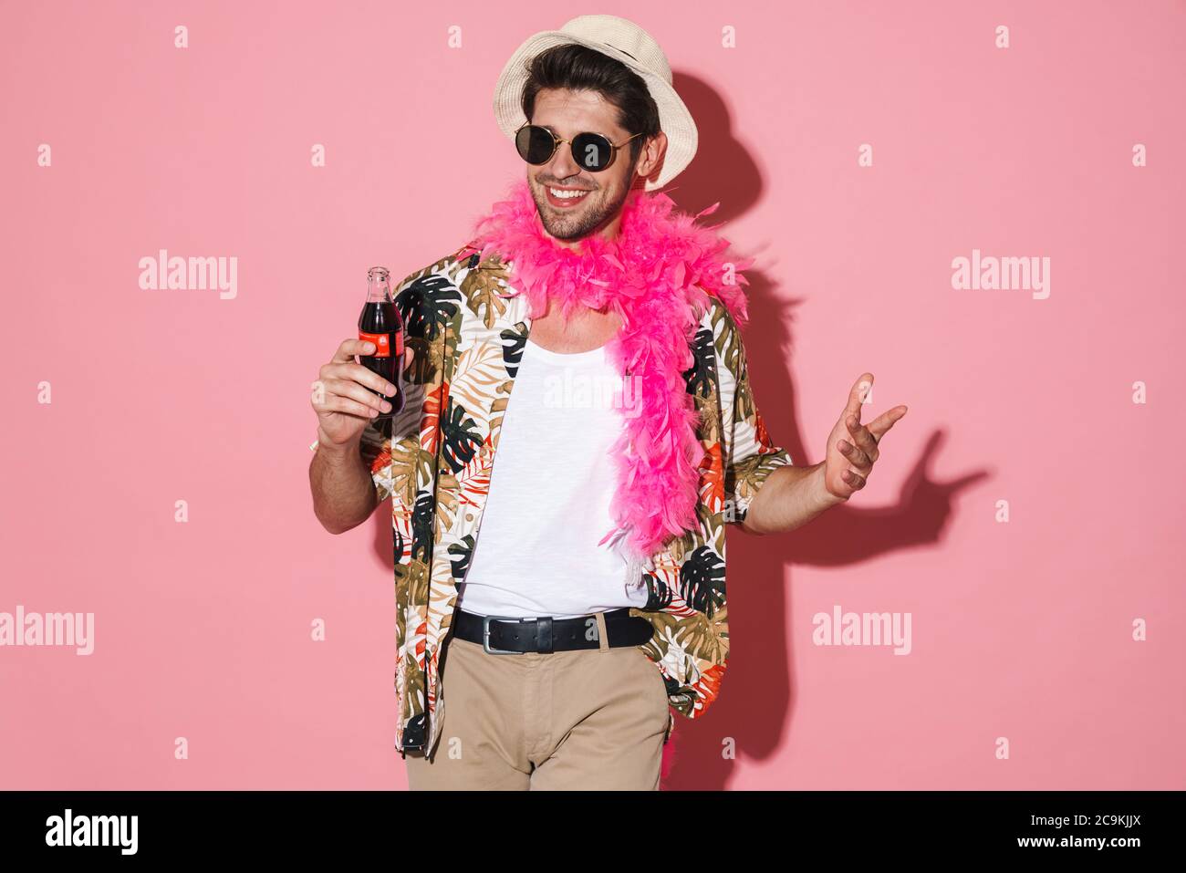 Portrait of cheerful young man wearing boa dancing while drinking soda ...