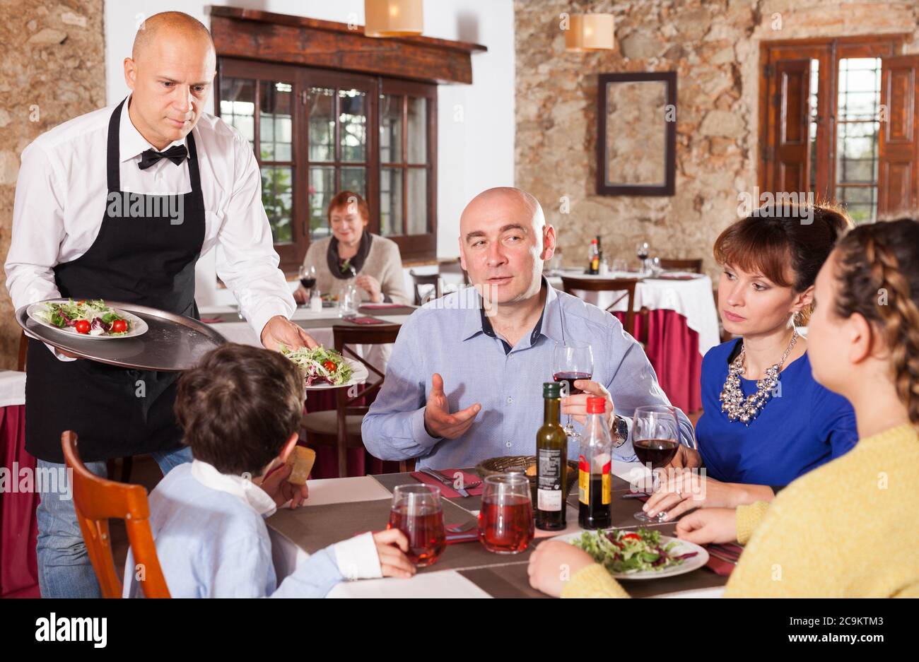 Cheerful waiter serving plates of salad to happy family having dinner ...