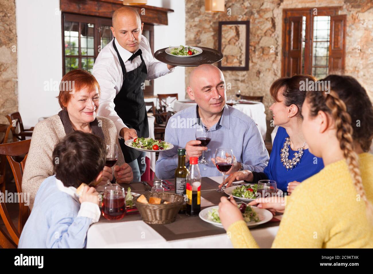 Cheerful waiter serving plates of salad to happy family having dinner ...