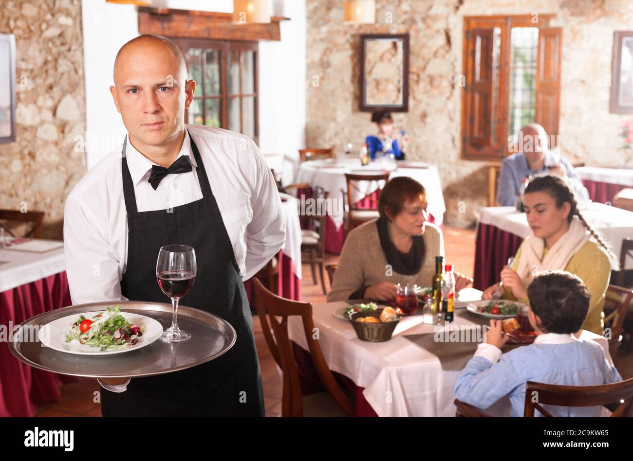 Positive waiter with tray in foreground and visitors having dinner at ...