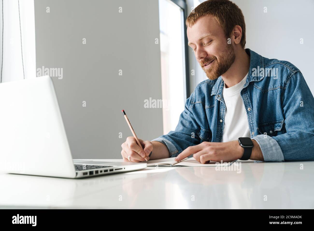 Photo of smiling handsome man writing down notes while working with ...