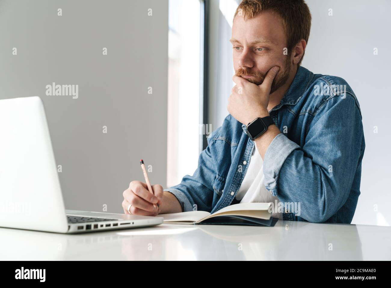 Photo of thinking handsome man writing down notes while working with ...