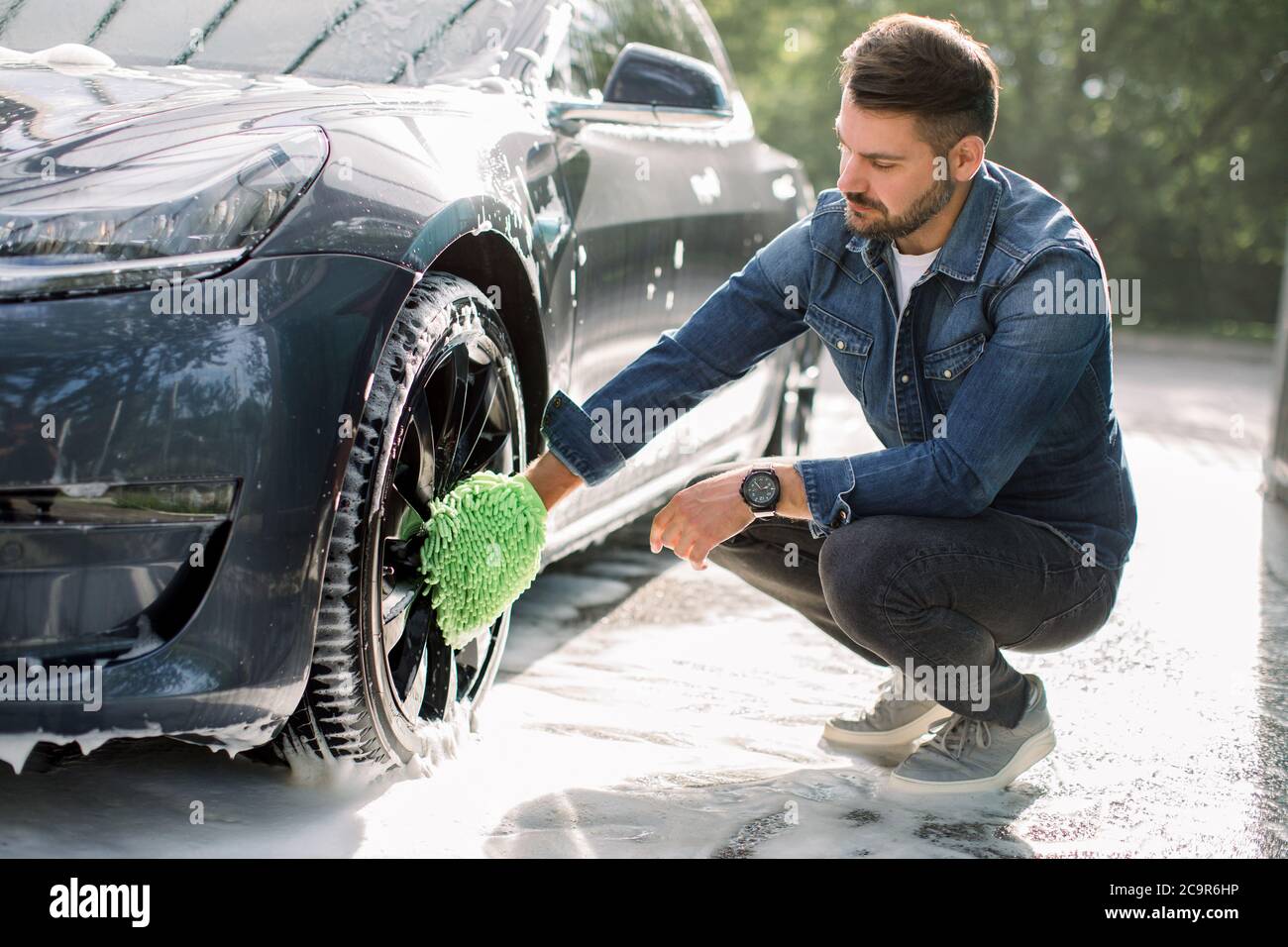 Young handsome bearded guy holding green sponge, washing car wheel with ...