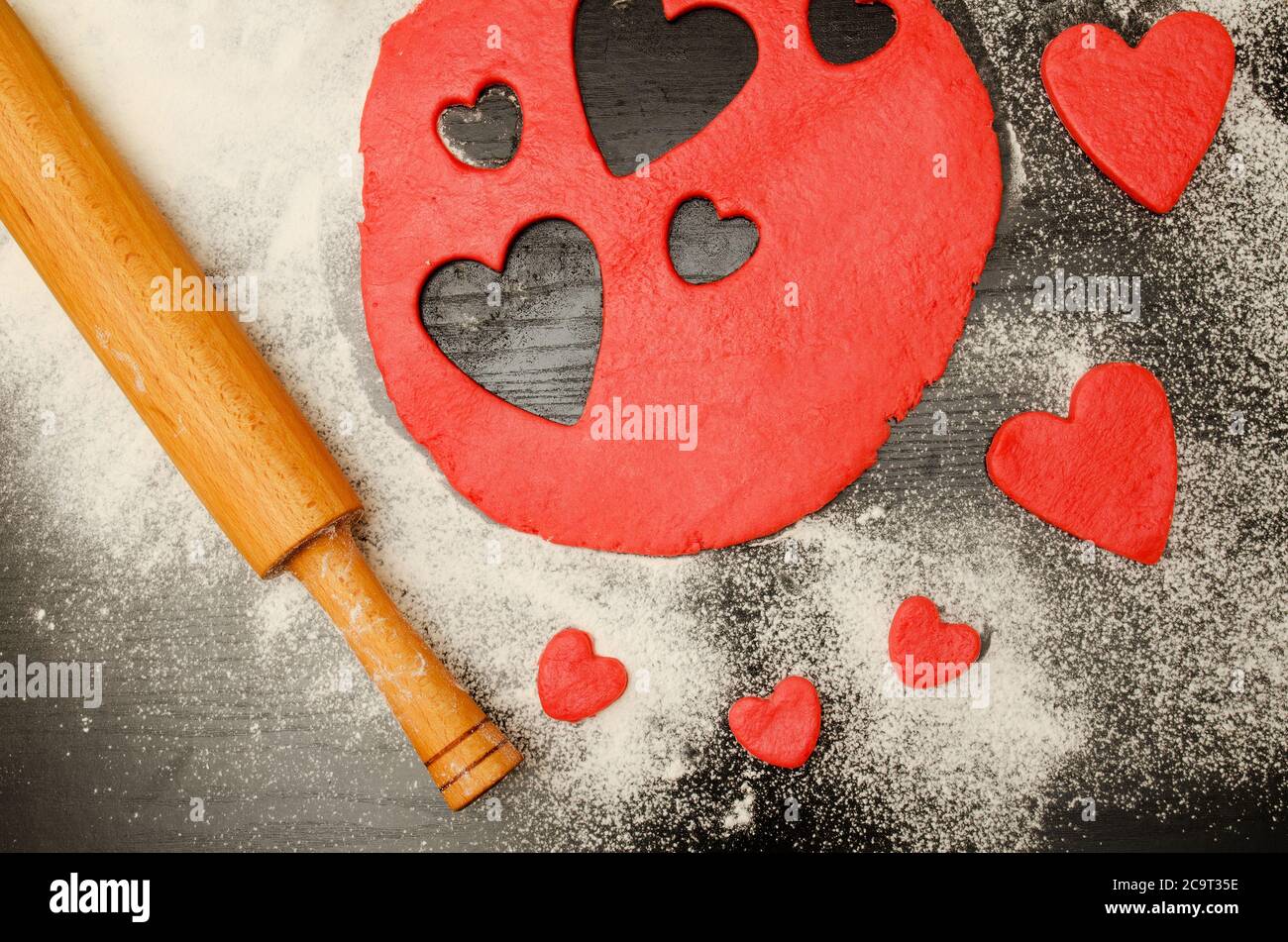 Red dough with cut-out hearts and flour on the black table, a rolling ...