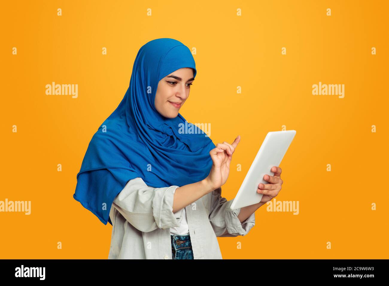 Holding tablet, scrolling. Young muslim woman on yellow studio ...
