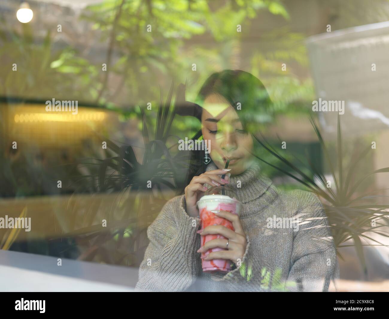 Portrait shot trough the window of young female drinking smoothie while ...