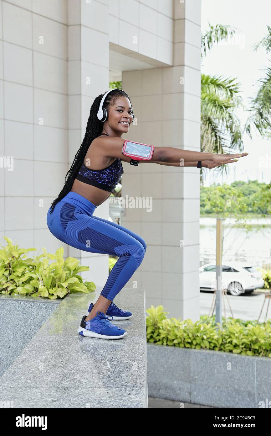 Smiling pretty young Black woman doing squats outdoors after jogging to ...