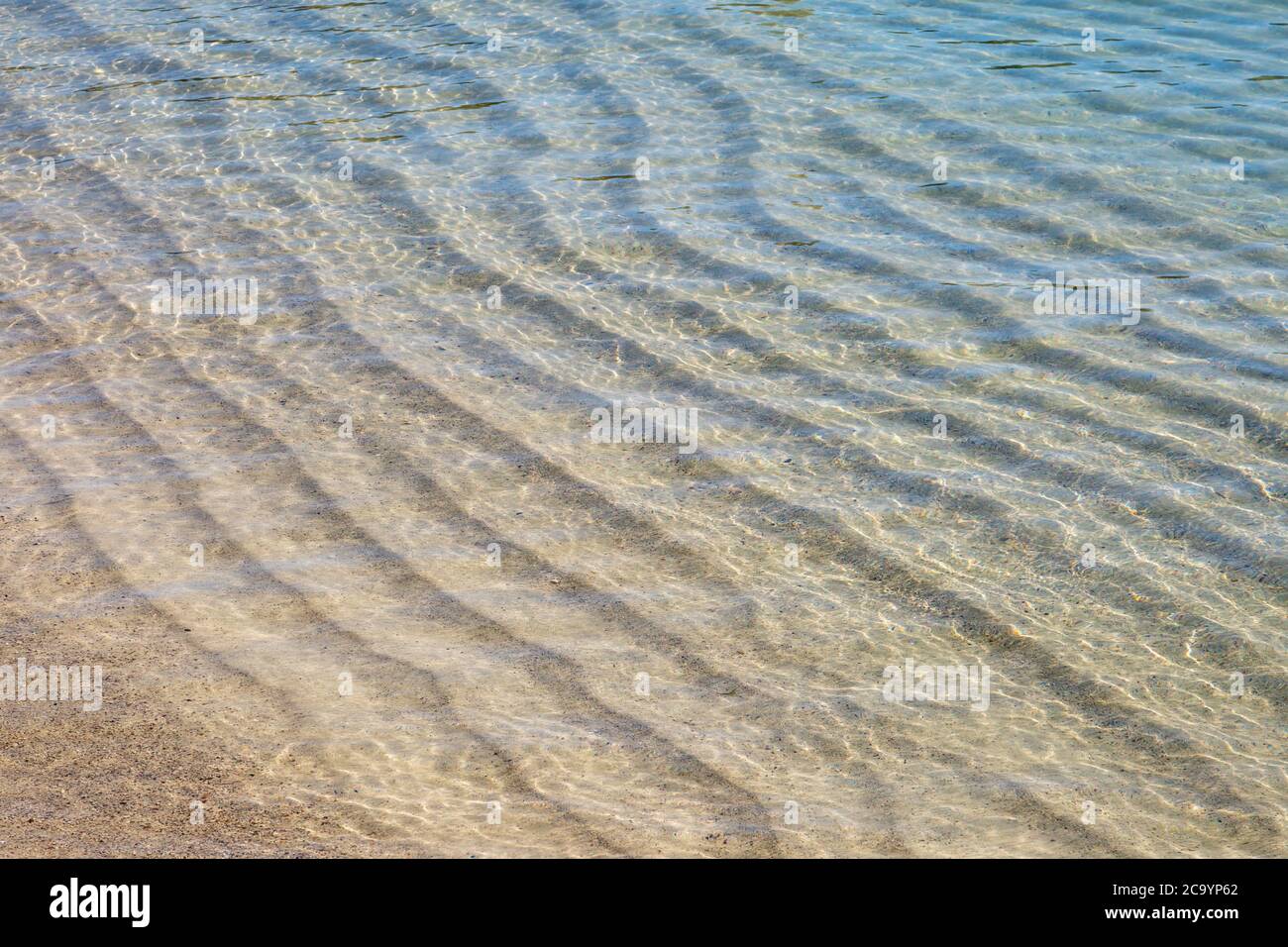 The shallow water of the ocean, with ripples in the sand visible below ...