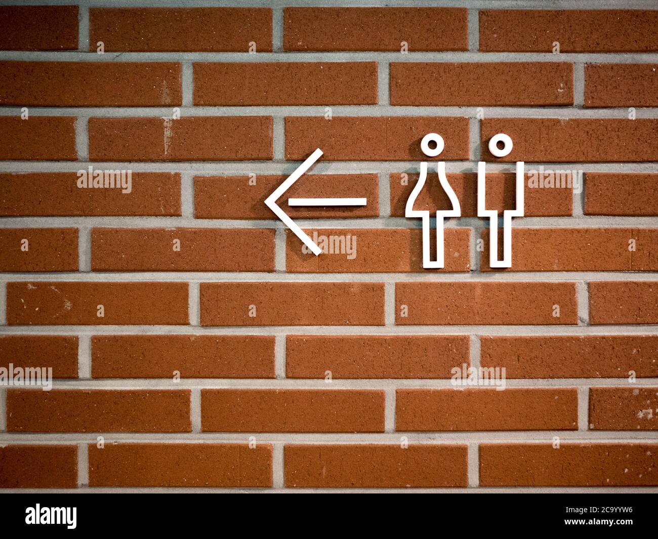 A directional sign to the toilet in a background of red bricks. A white ...