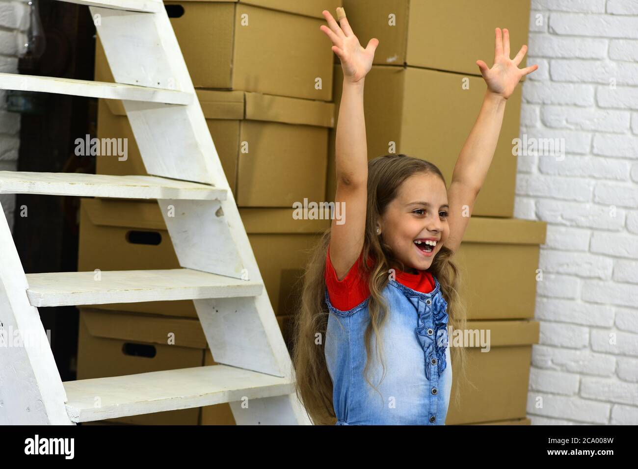 Kid stands by pile of cardboard boxes and white ladder. Girl with fair ...
