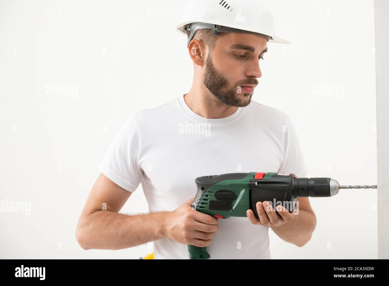 Concentrated young bearded man in construction hardhat drilling hole ...