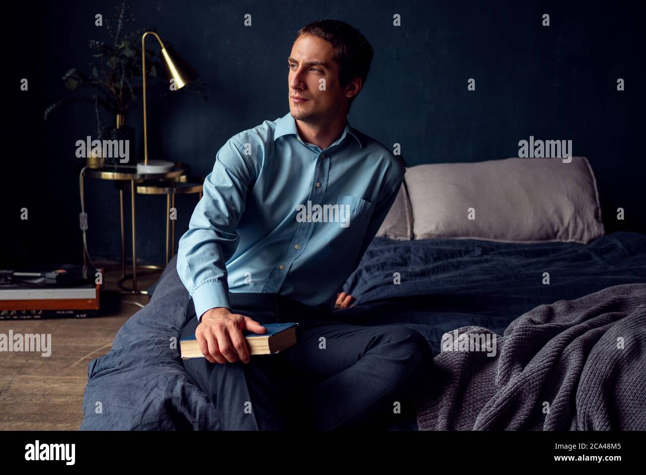Young man with a book is sitting on the bed, a portrait in a dark blue ...