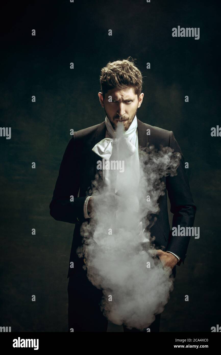 Vaping, trendy look. Young man in suit as Dorian Gray isolated on dark ...