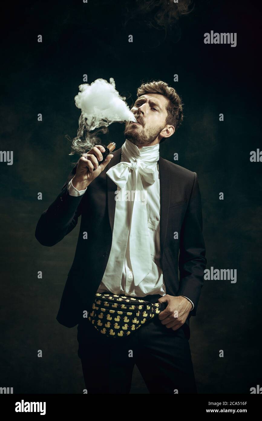 Vaping with smoke. Young man in suit as Dorian Gray isolated on dark ...