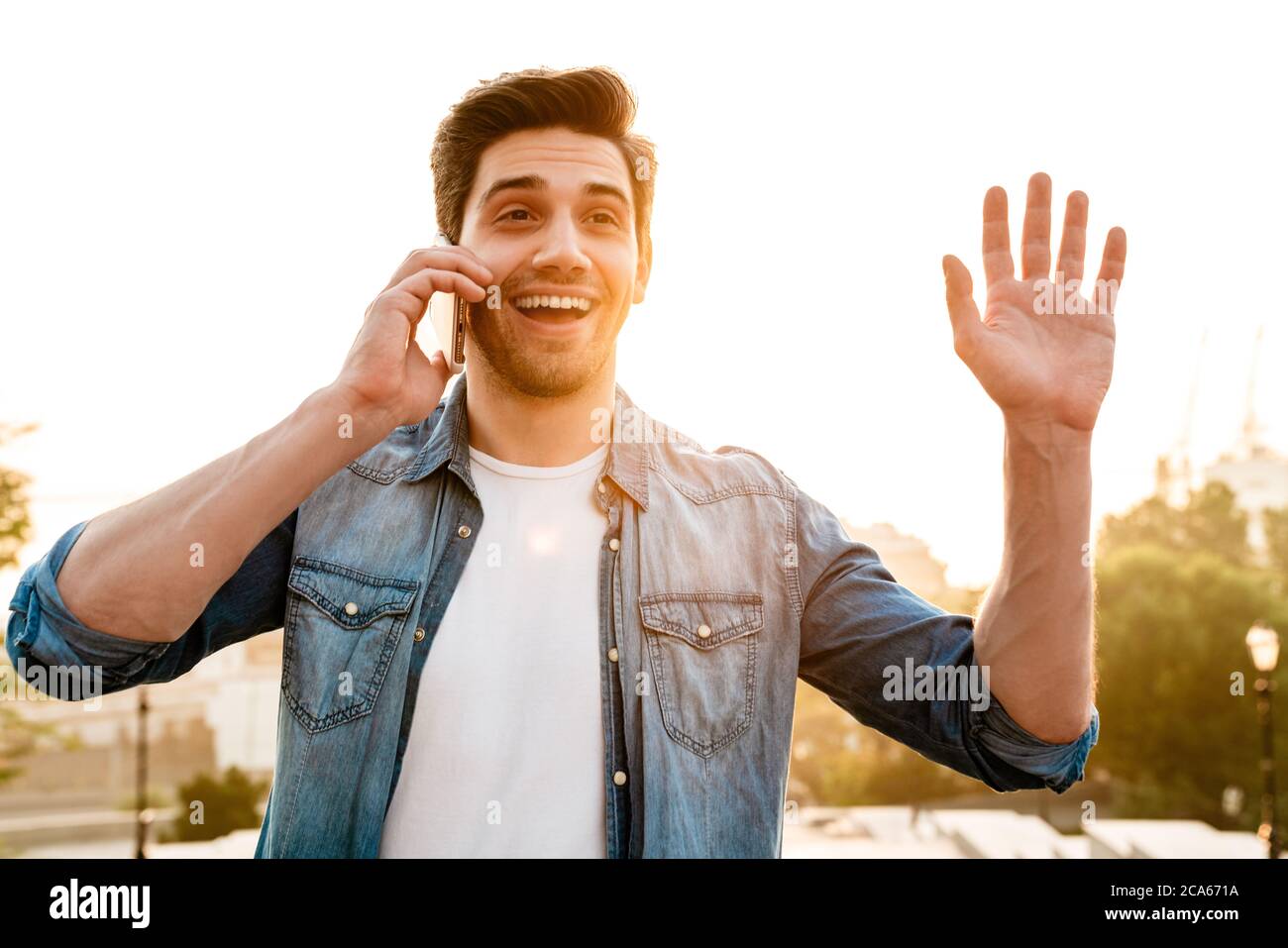 Photo of excited handsome man waving hand and talking on mobile phone ...