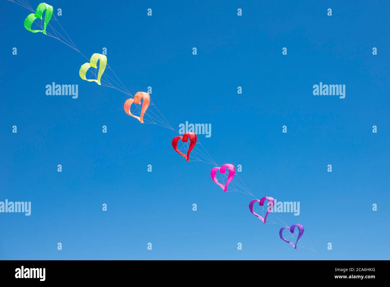 A row of rainbow coloured heart shaped kites flying in the blue sky ...