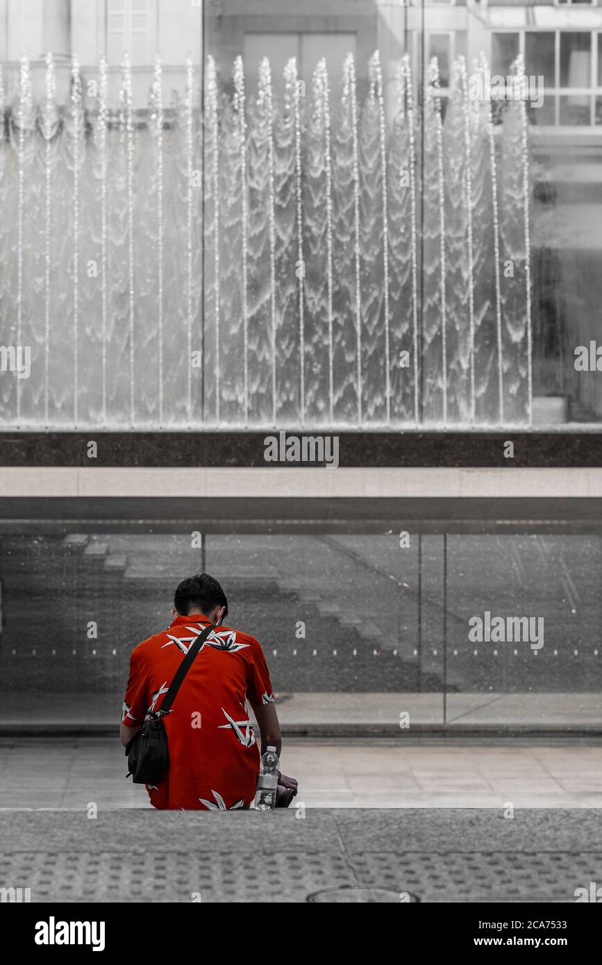 A man in a red shirt sits in front of a fountain, image of street ...