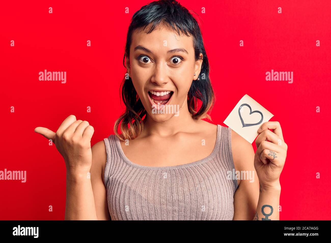Young woman holding heart reminder pointing thumb up to the side ...