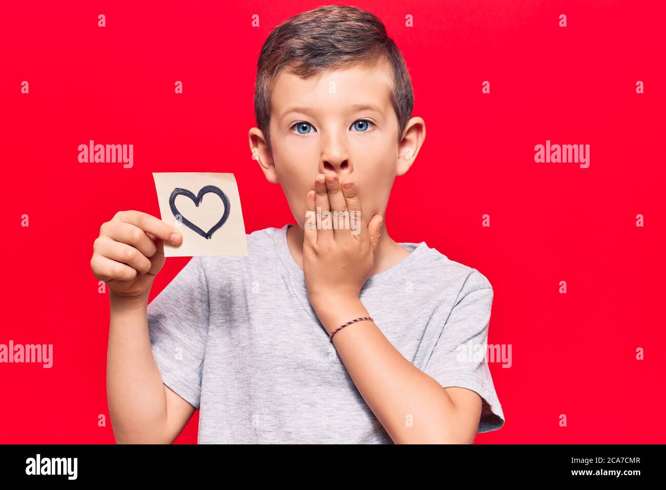 Cute blond kid holding heart reminder covering mouth with hand, shocked ...