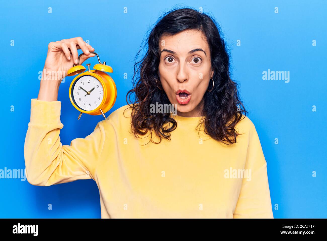 Young beautiful hispanic woman holding alarm clock scared and amazed ...