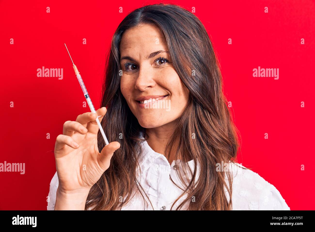 Young beautiful brunette woman holding syringe over isolated red ...