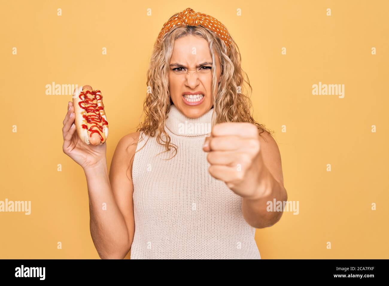 Young beautiful blonde woman eating fast food hot dog with ketchup and ...