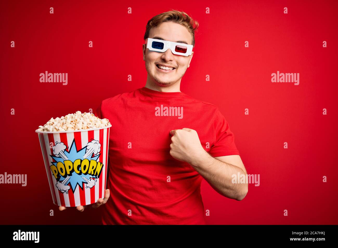 Young handsome redhead man watching 3d glasses eating popcorn snack over red background with ...