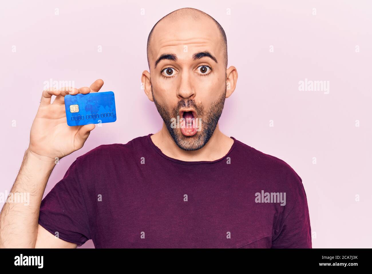Young handsome bald man holding credit card scared and amazed with open ...
