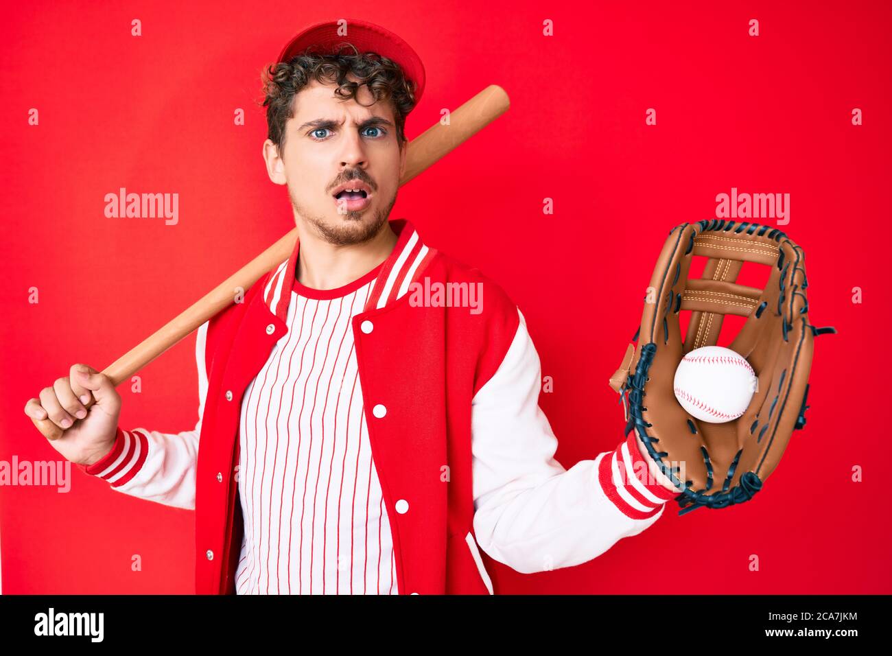 Young caucasian man with curly hair wearing baseball uniform holding ...